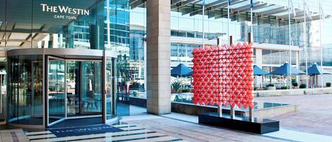 Entrada de hotel moderna com portas de vidro, uma escultura geométrica vermelha e um calçadão ladrilhado.