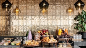 Buffet de café da manhã com diversas opções de alimentos e bebidas, balcão de bancada de mármore e luminárias pendentes.