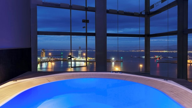 Piscina interna moderna com vista panorâmica de uma paisagem urbana ao entardecer.