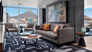 Uma sala de estar moderna com sofá, poltronas e mesa de centro. Um grande quadro abstrato está na parede, e há vista para um horizonte urbano e uma montanha ao fundo.