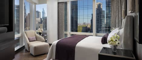 Quarto de hotel moderno com uma cama grande, uma poltrona, uma mesinha de cabeceira e vista para o horizonte urbano através de amplas janelas.