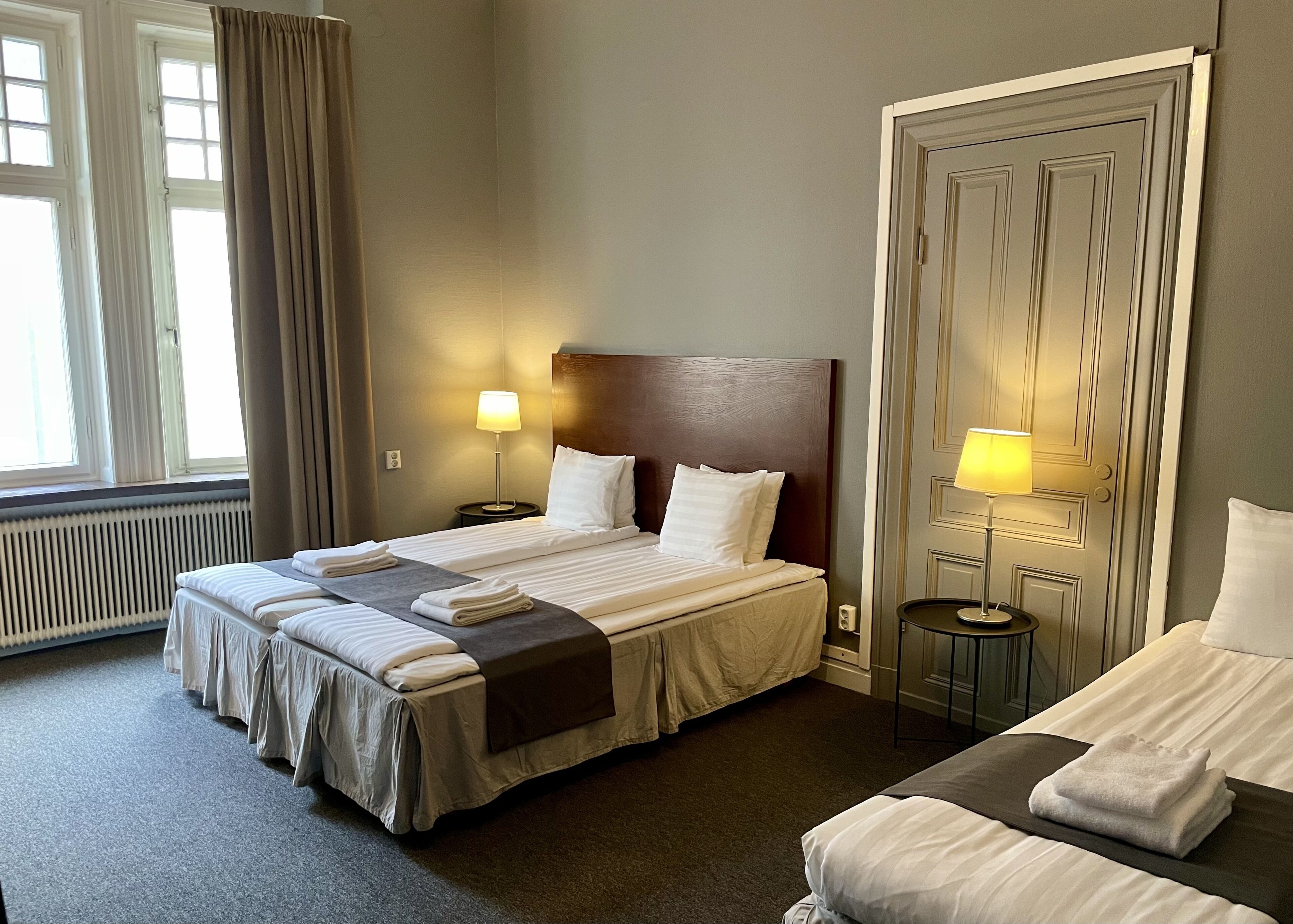 Quarto de hotel com uma cama grande, duas mesas de cabeceira, um criado-mudo, uma janela com cortinas e uma porta com painel decorativo.