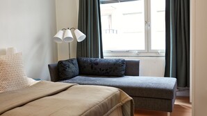 Quarto com cama, sofá cinza e janela com cortinas.