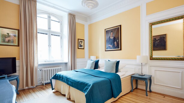 Quarto com cama, mesa de cabeceira, televisão, janela com cortinas e quadros na parede.