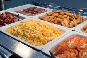 Buffet com diversas opções de comida, incluindo bacon, ovos, batatas fritas e tomates.