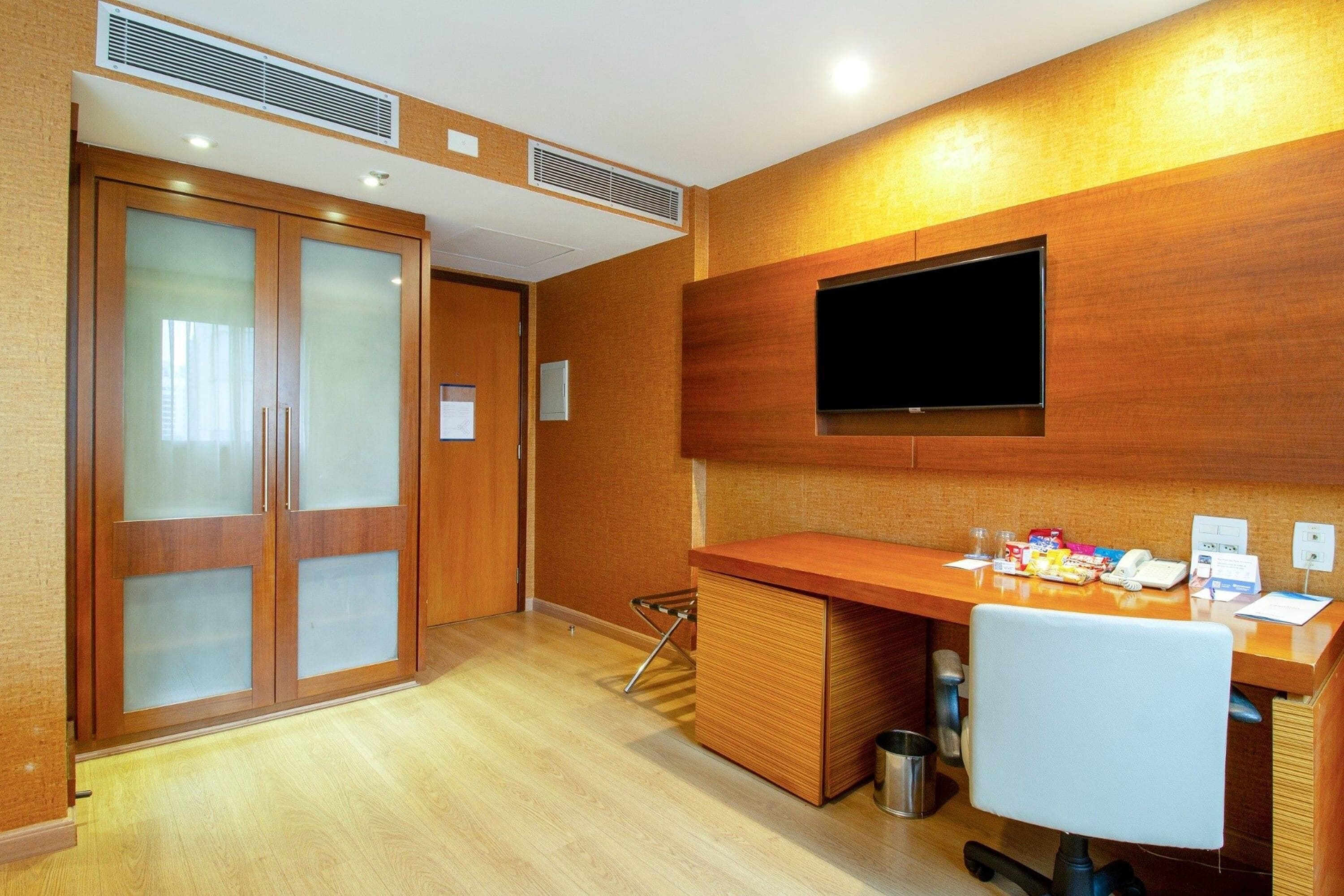 Quarto de hotel com uma secretária de madeira, uma cadeira, uma televisão fixada na parede, uma porta de correr com painéis de vidro jateado e uma lixeira.