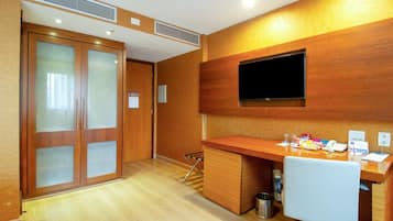 Quarto de hotel com uma secretária de madeira, uma cadeira, uma televisão fixada na parede, uma porta de correr com painéis de vidro jateado e uma lixeira.