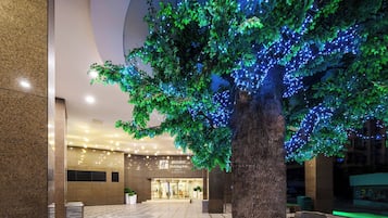 Uma árvore com luzes azuis, entrada de um prédio e piso de azulejos.