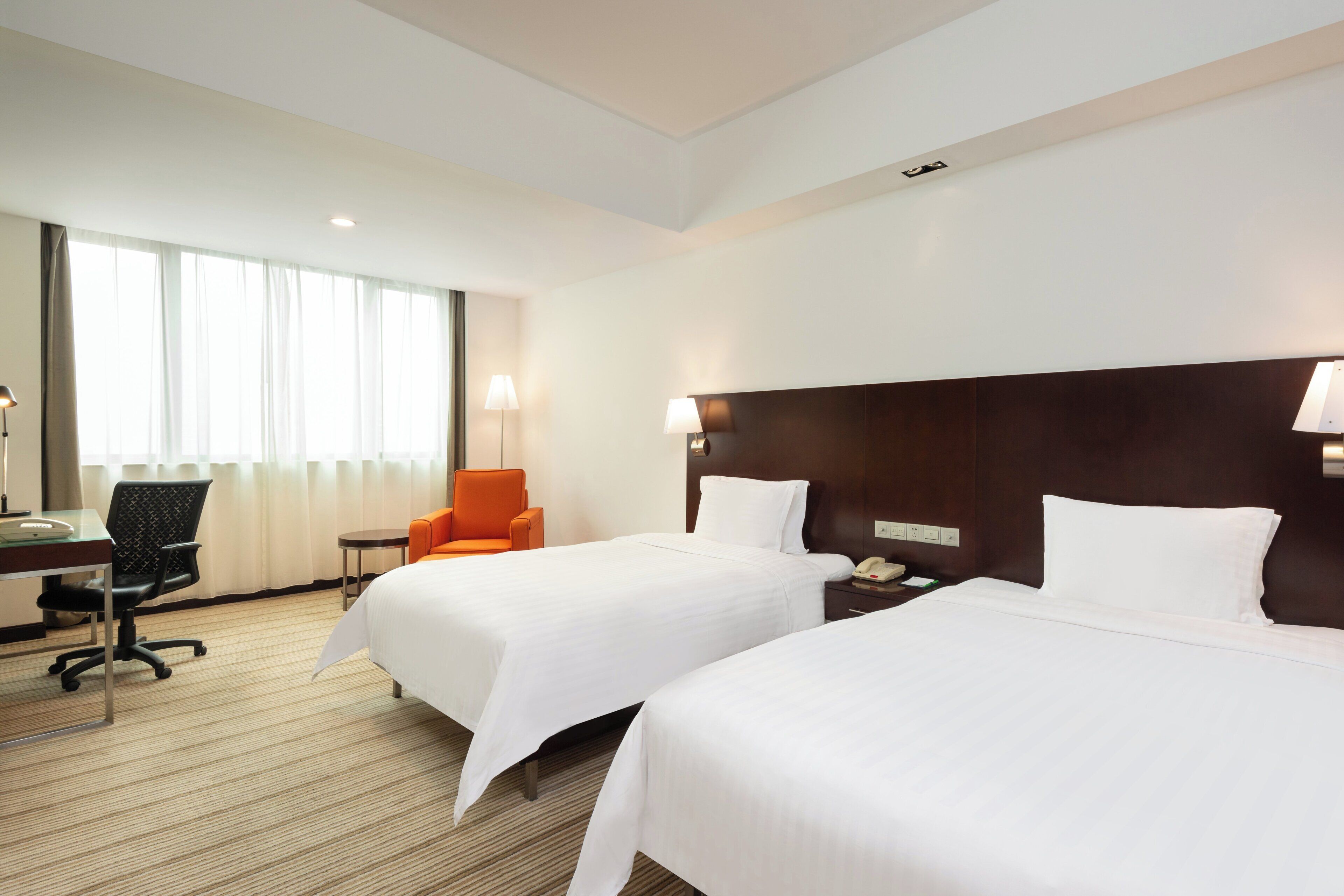 Quarto de hotel com duas camas, uma escrivaninha, uma cadeira laranja e uma janela com cortinas.