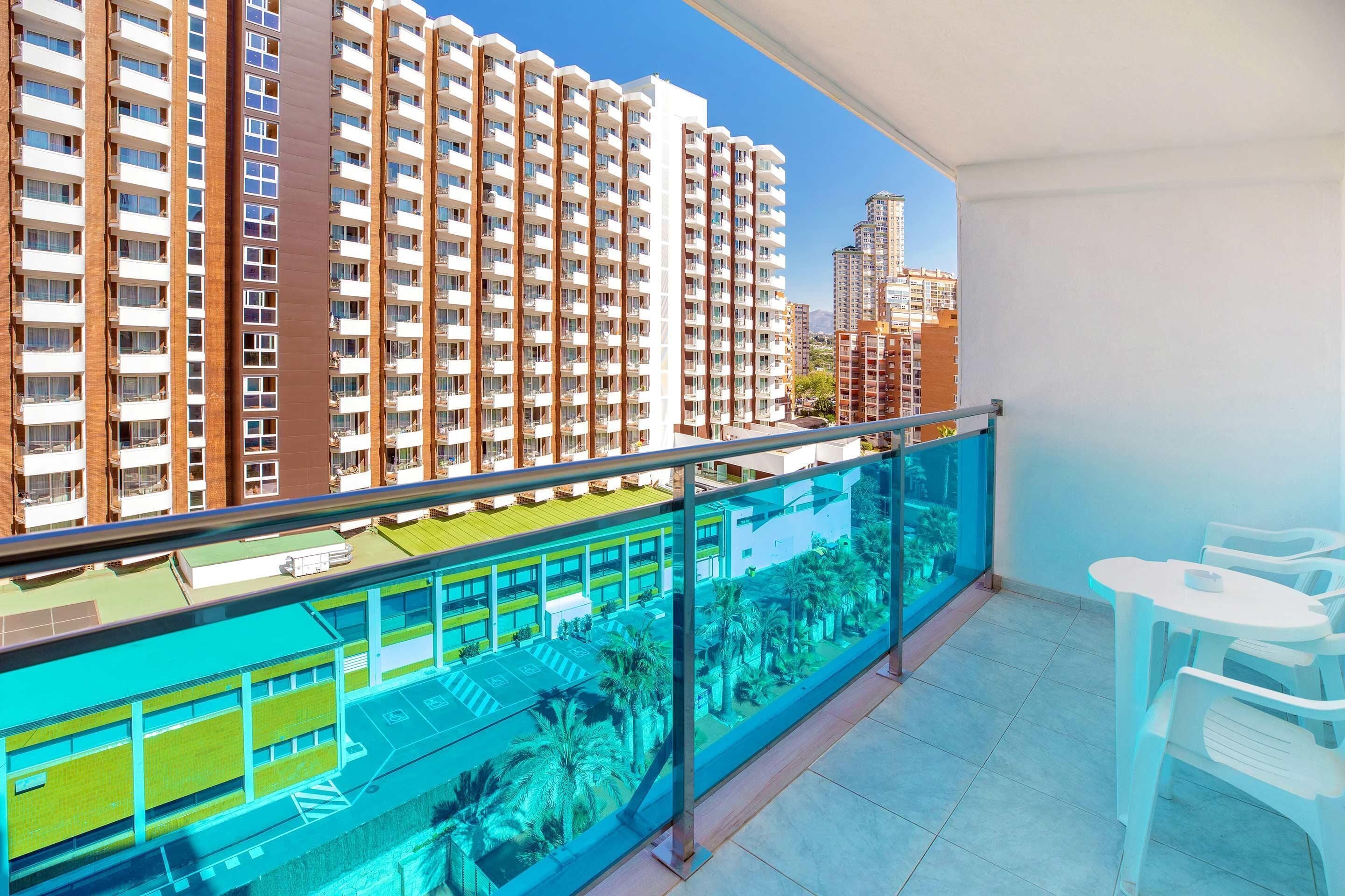 Varanda com vista para edifícios altos, guarda-corpo de vidro e cadeiras de plástico branco.