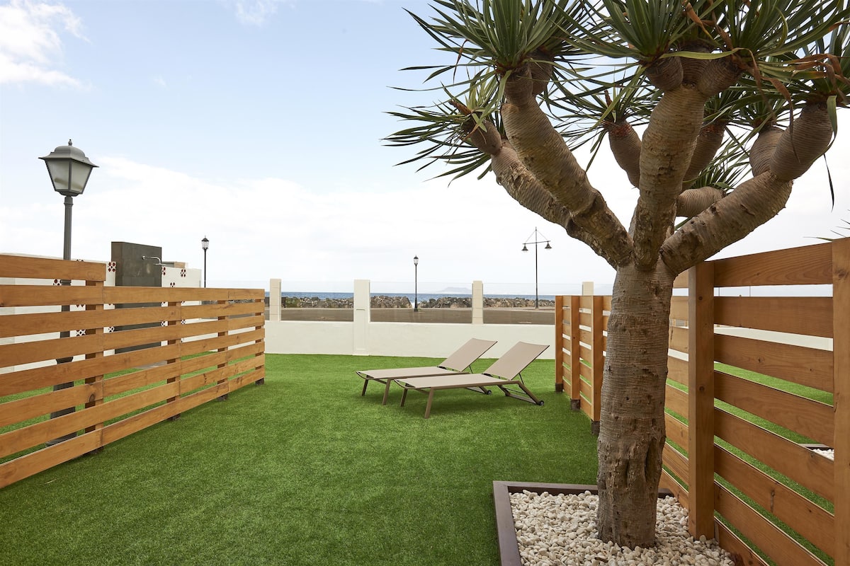 Terraço no telhado com grama sintética, duas cadeiras de descanso, cerca de madeira e uma palmeira.