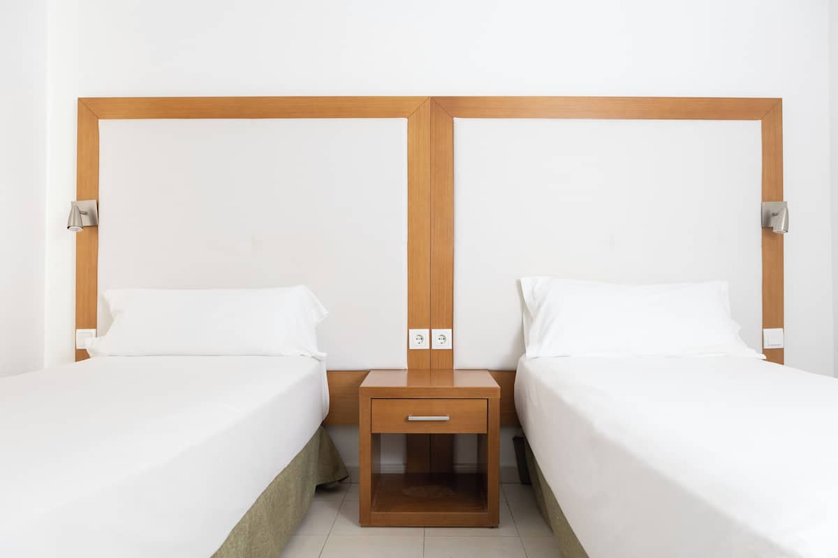 Quarto de hotel com duas camas de solteiro, uma mesa de cabeceira de madeira com gaveta e luminária fixa na parede.