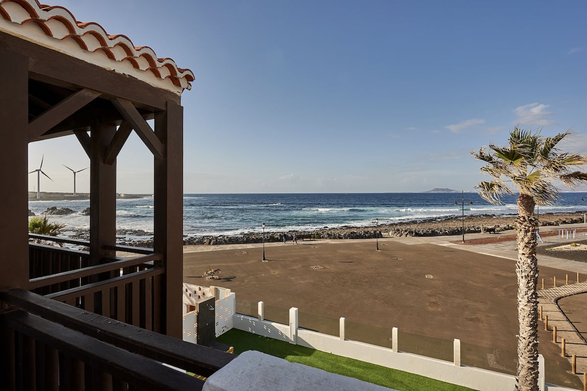 Vista da praia a partir de uma área coberta com deck de madeira, uma palmeira e turbinas eólicas ao fundo.