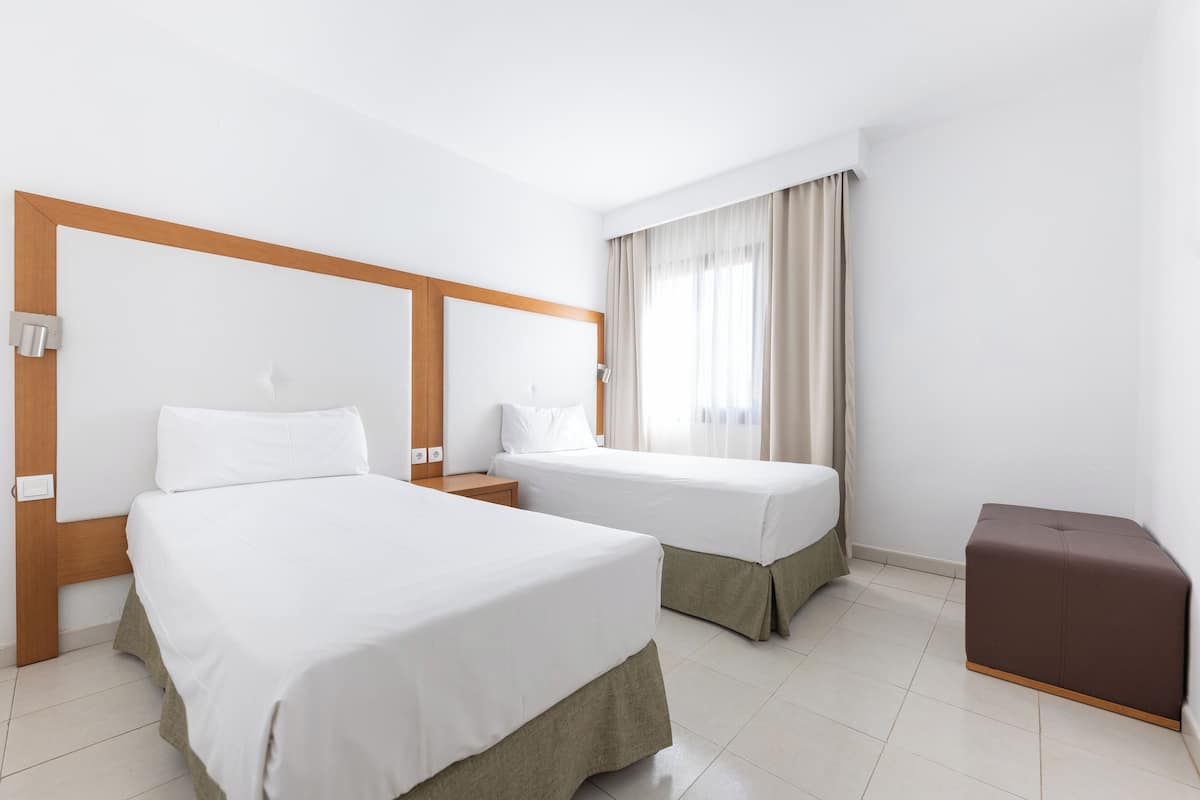 Quarto de hotel com duas camas, cabeceira de madeira, um banquinho marrom e uma janela com cortinas.