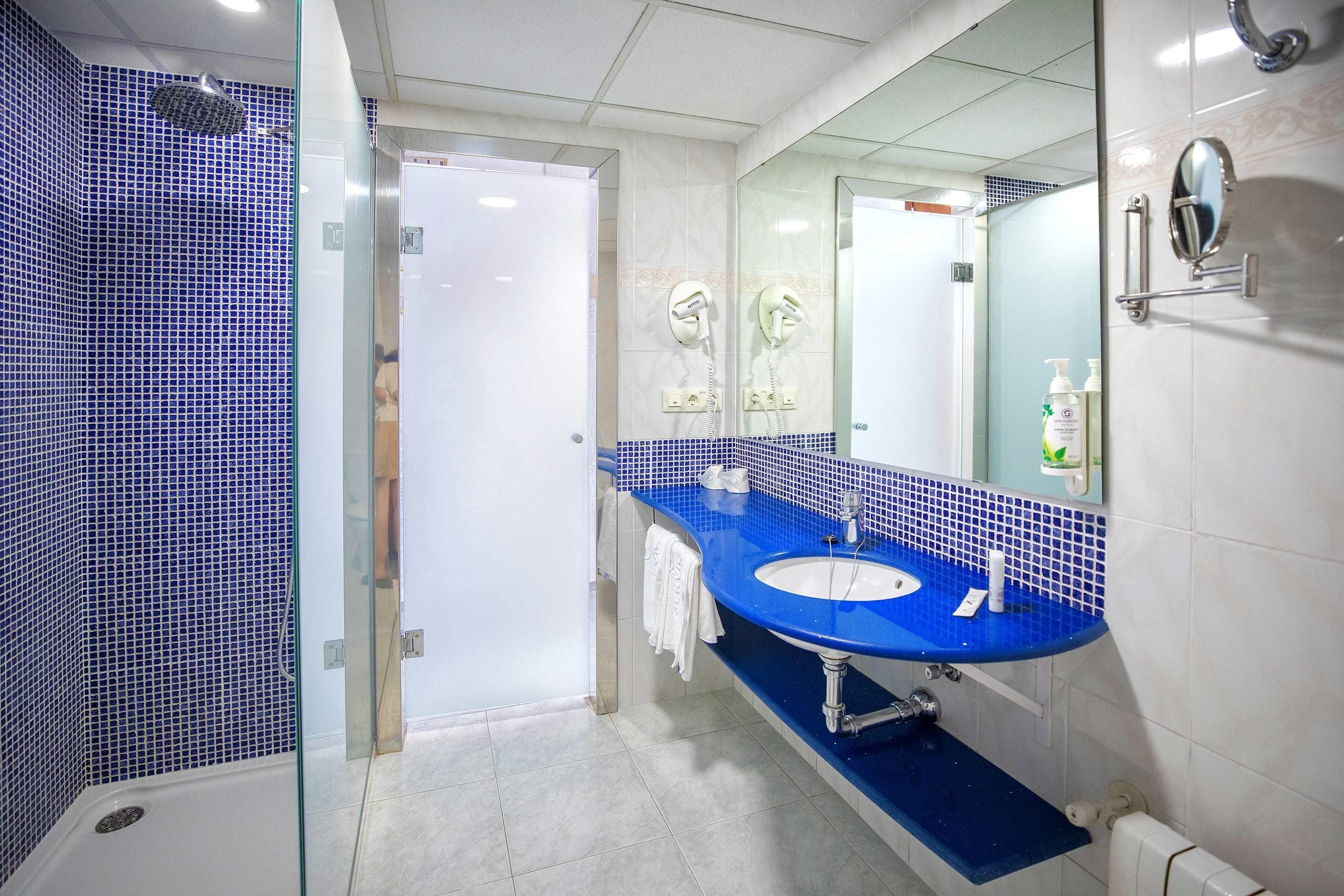 Um banheiro com área de chuveiro em azulejos azuis, pia branca com bancada azul e espelho com toalha pendurada na parede.