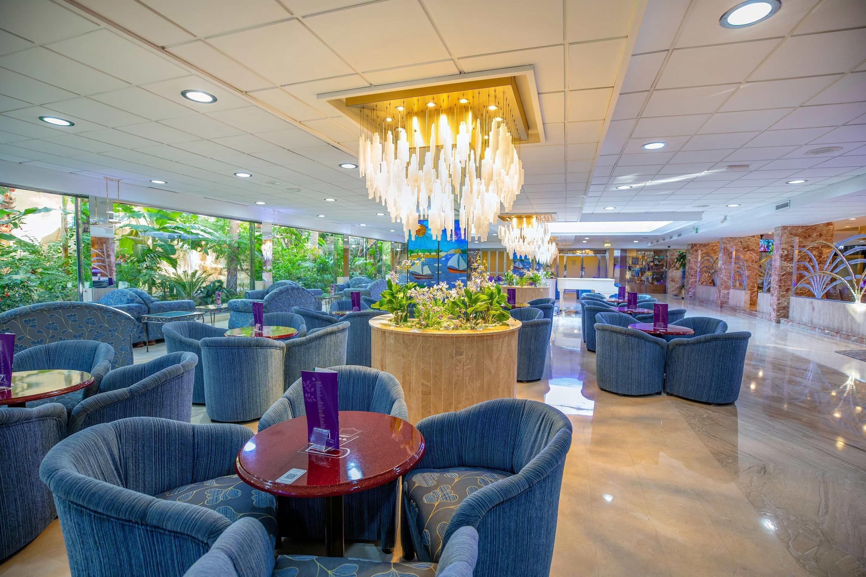 Um saguão de hotel moderno com assentos estofados em azul, uma mesa redonda central e um lustre decorativo.