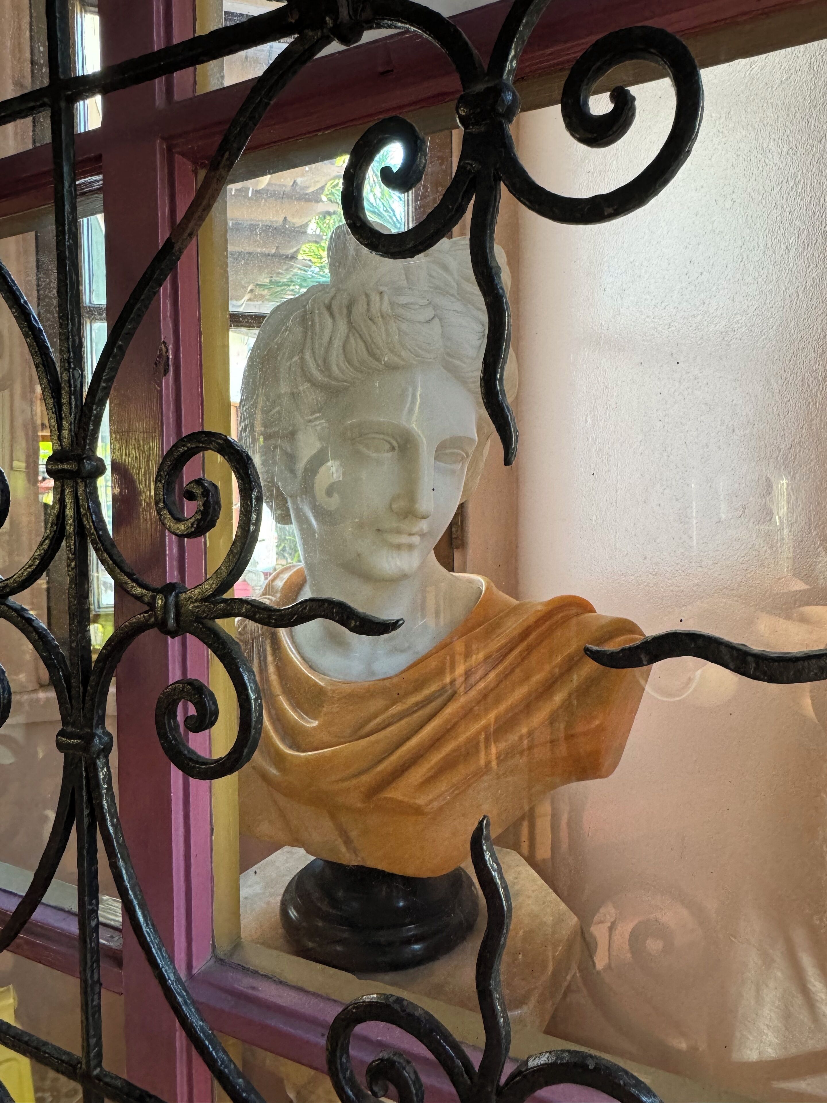 Escultura de busto com vestimenta drapeada, diante de uma parede com grades de ferro decorativas.