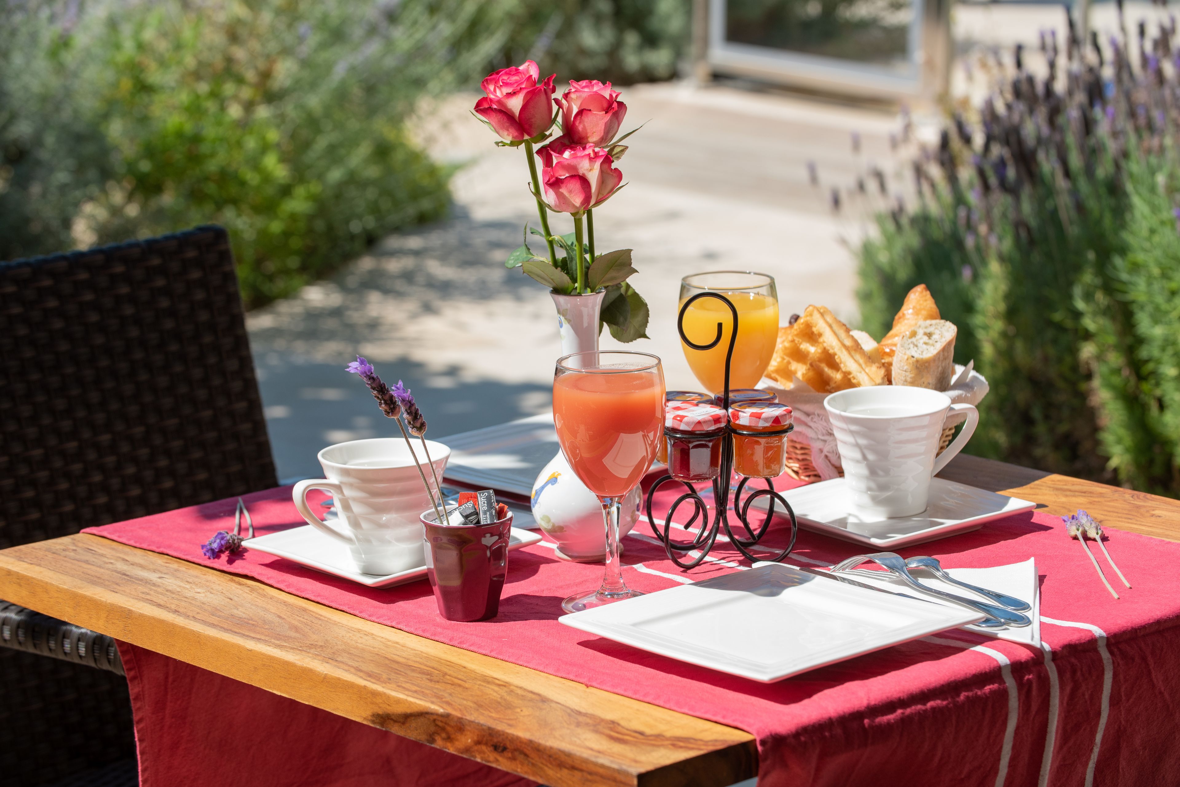 Mesa de café da manhã com toalha vermelha, um vaso com rosas cor-de-rosa, um copo de suco de laranja, uma xícara de café, um prato com torradas e um pote pequeno de geleia.