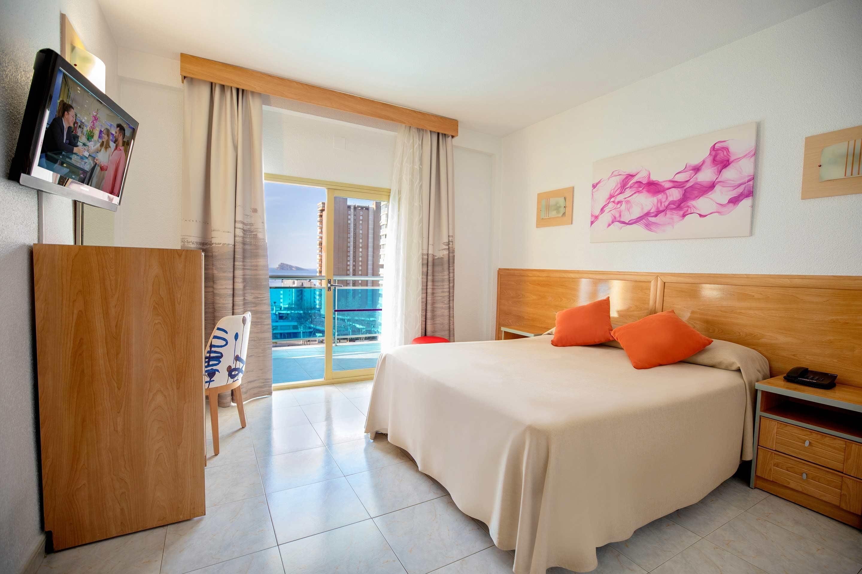 Quarto de hotel com cama, escrivaninha, cadeira, televisão e varanda com vista para edifícios.