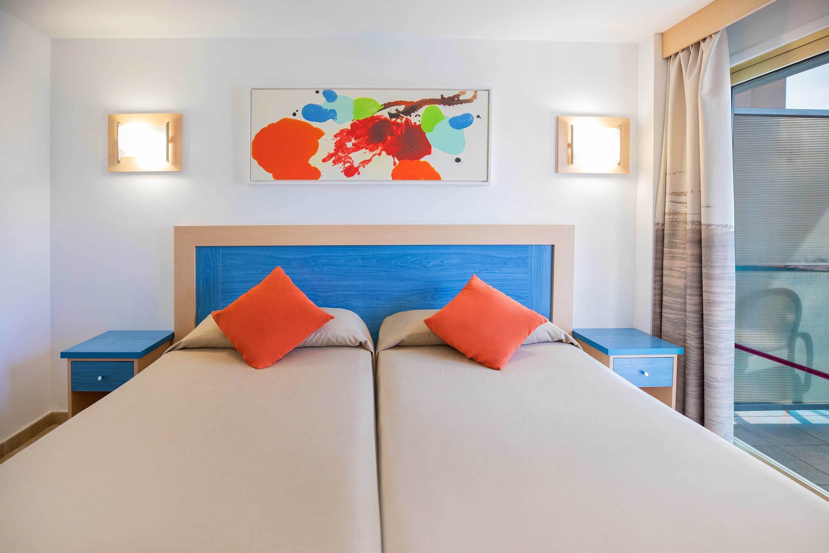 Uma cama de casal com duas almofadas laranjas, um painel de cabeceira azul e um quadro abstrato emoldurado acima.
