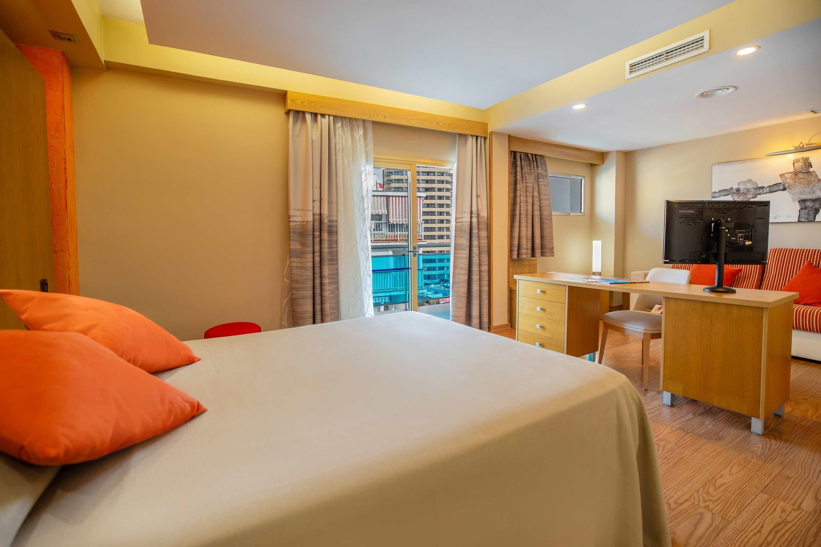 Quarto de hotel com cama, escrivaninha, cadeira e televisão.