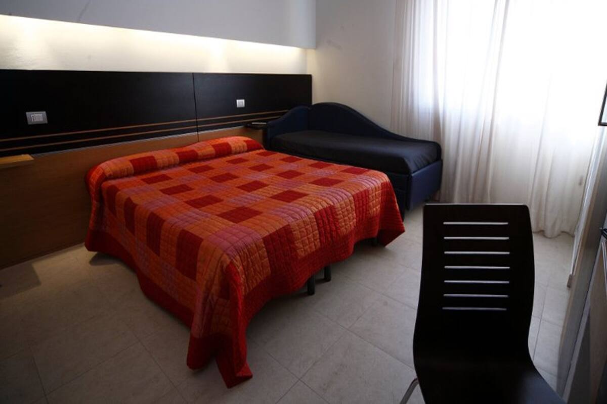 Quarto de hotel com uma cama, uma cadeira e uma janela com cortinas.
