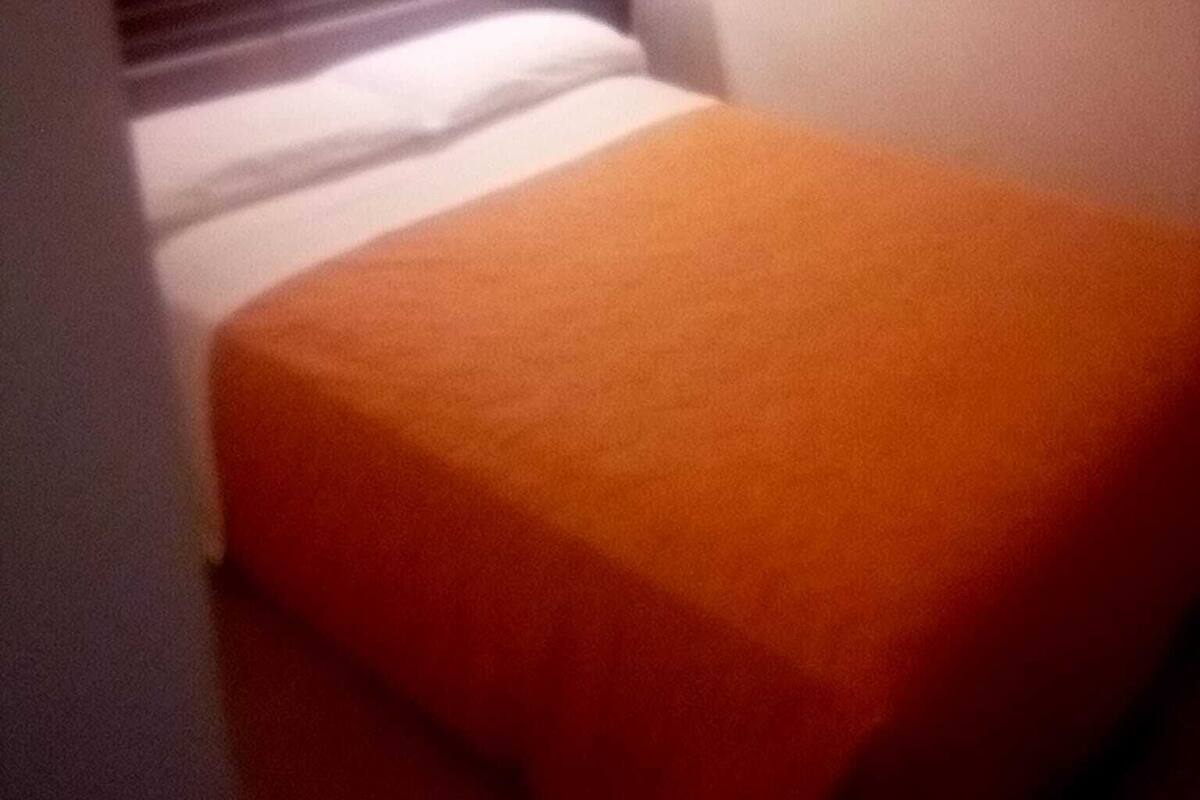 Uma cama de solteiro com colcha laranja num quarto com parede branca e cabeceira de madeira.