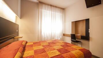 Quarto de hotel com uma cama grande, uma TV de tela plana na parede, uma escrivaninha com uma cadeira e uma janela com cortinas finas.