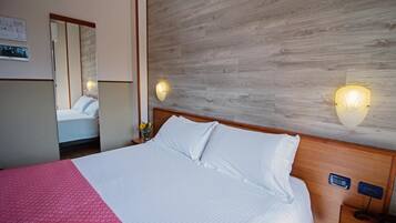 Quarto de hotel com cabeceira de madeira, cama com roupa de cama branca, mesa de cabeceira com telefone e luminária fixada na parede.