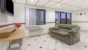 Quarto com sofá verde, mesa de madeira, televisão de tela plana, janela com persianas e piso de azulejos.