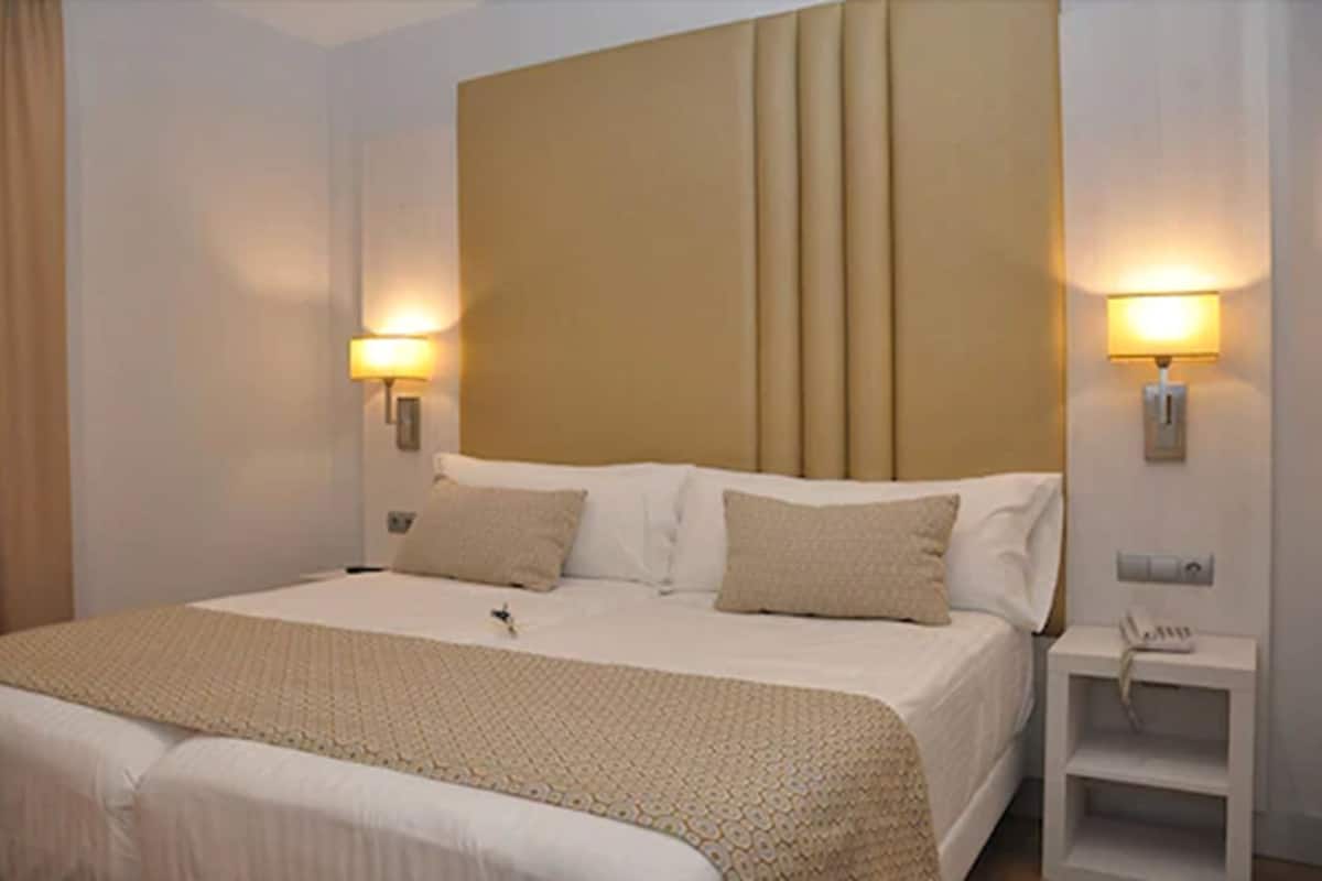 Quarto de hotel com uma cama grande, duas luminárias de cabeceira, mesa de cabeceira com telefone e cabeceira com painéis verticais.