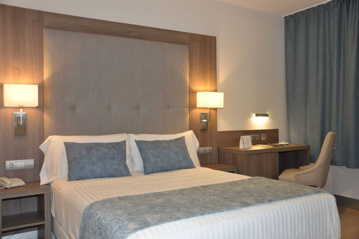 Quarto de hotel com uma cama grande, duas luminárias de cabeceira, uma escrivaninha com cadeira e um telefone na mesinha de cabeceira.