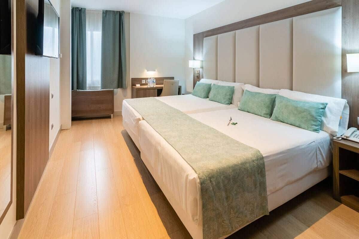 Quarto de hotel com cama grande, mesas de cabeceira, escrivaninha, televisão e janela com cortinas.