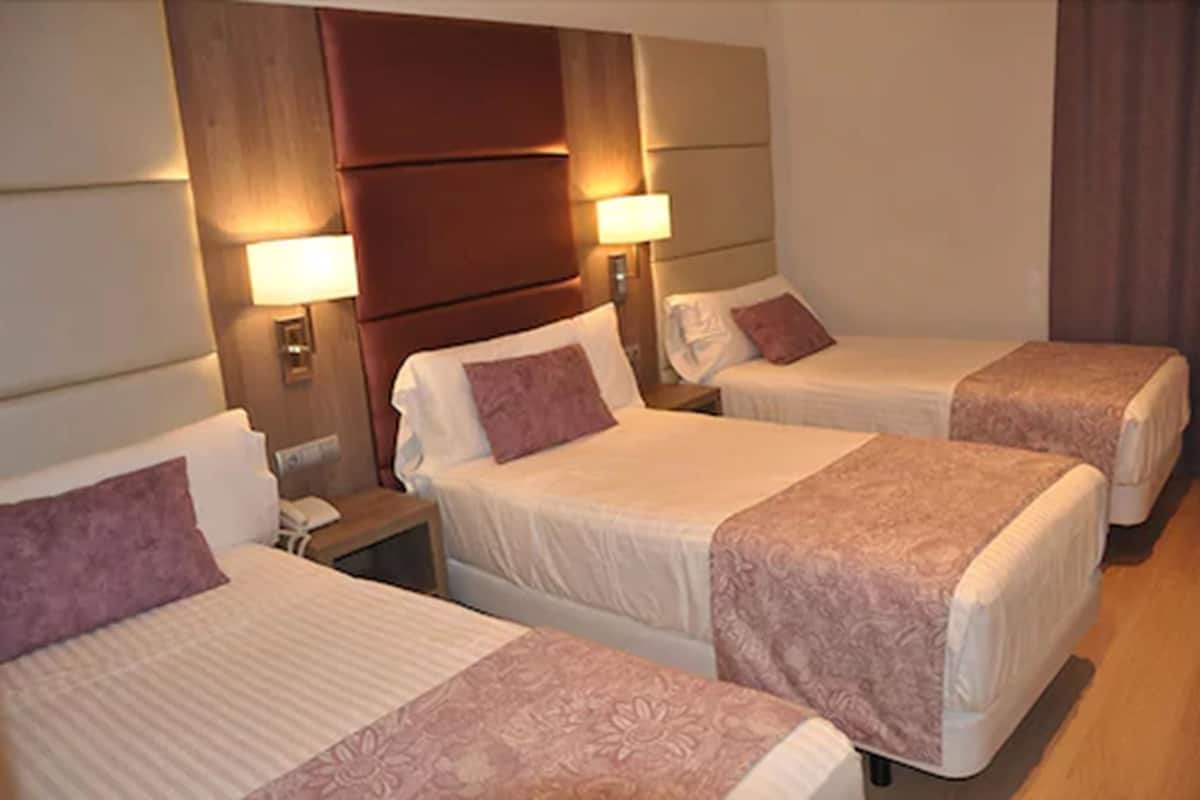 Quarto de hotel com três camas de solteiro, cada uma com cabeceira, mesas de cabeceira e luminárias.