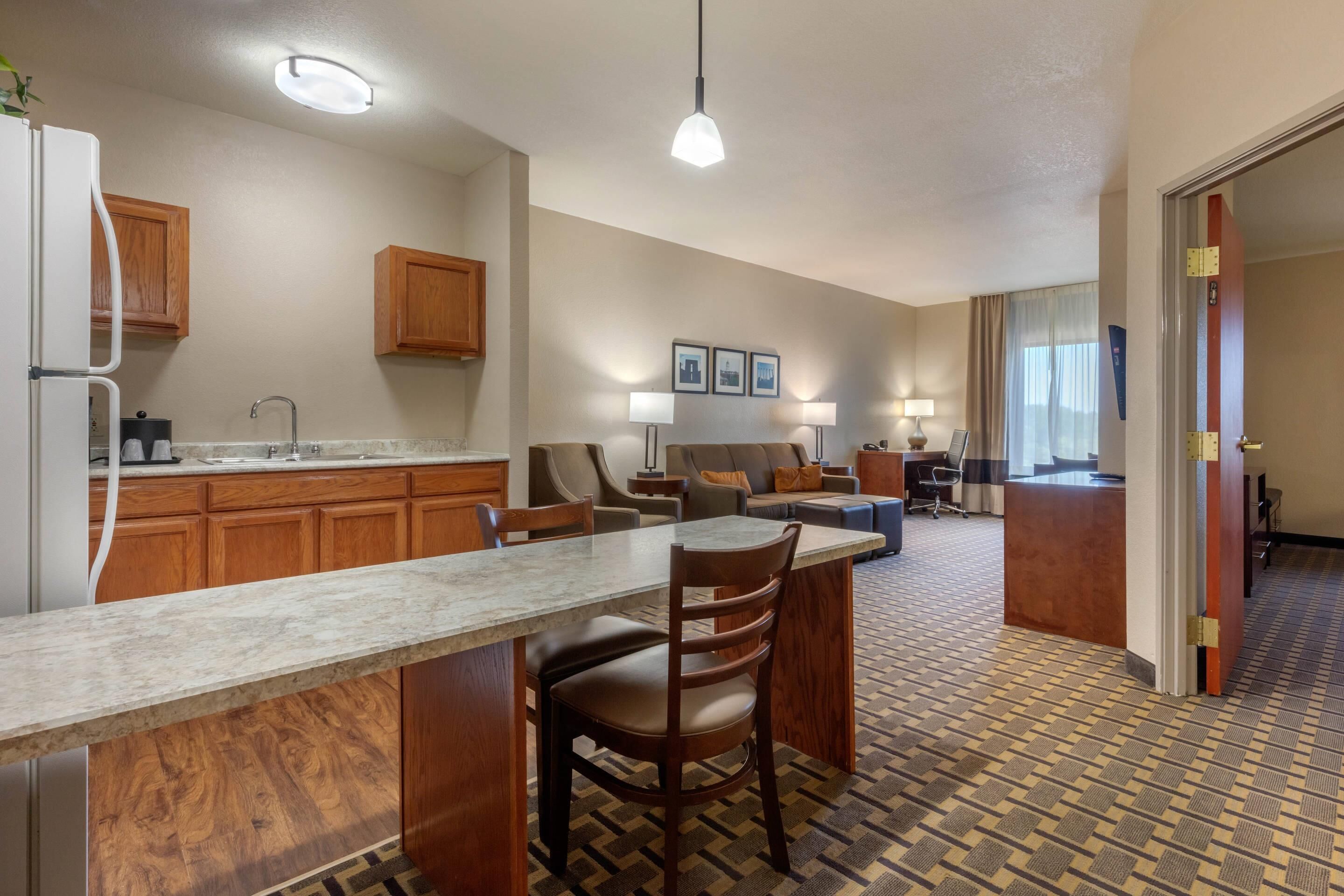 Quarto de hotel com cozinha compacta, área de refeições, sala de estar com sofá e televisão, e um tapete estampado.