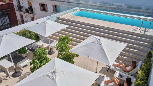 Uma área de piscina no terraço, com guarda-sóis brancos, espreguiçadeiras e degraus que dão acesso à piscina.