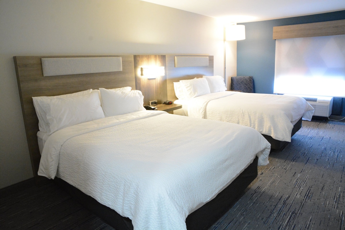 Quarto de hotel com duas camas, uma mesa de cabeceira, uma luminária e uma janela com persianas.
