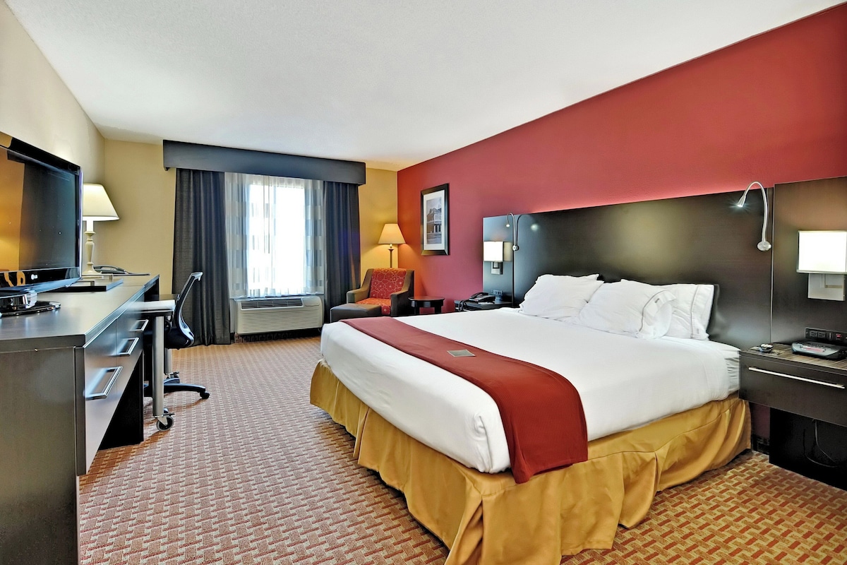 Quarto de hotel com cama grande, escrivaninha, cadeira, televisão e janela com cortinas.