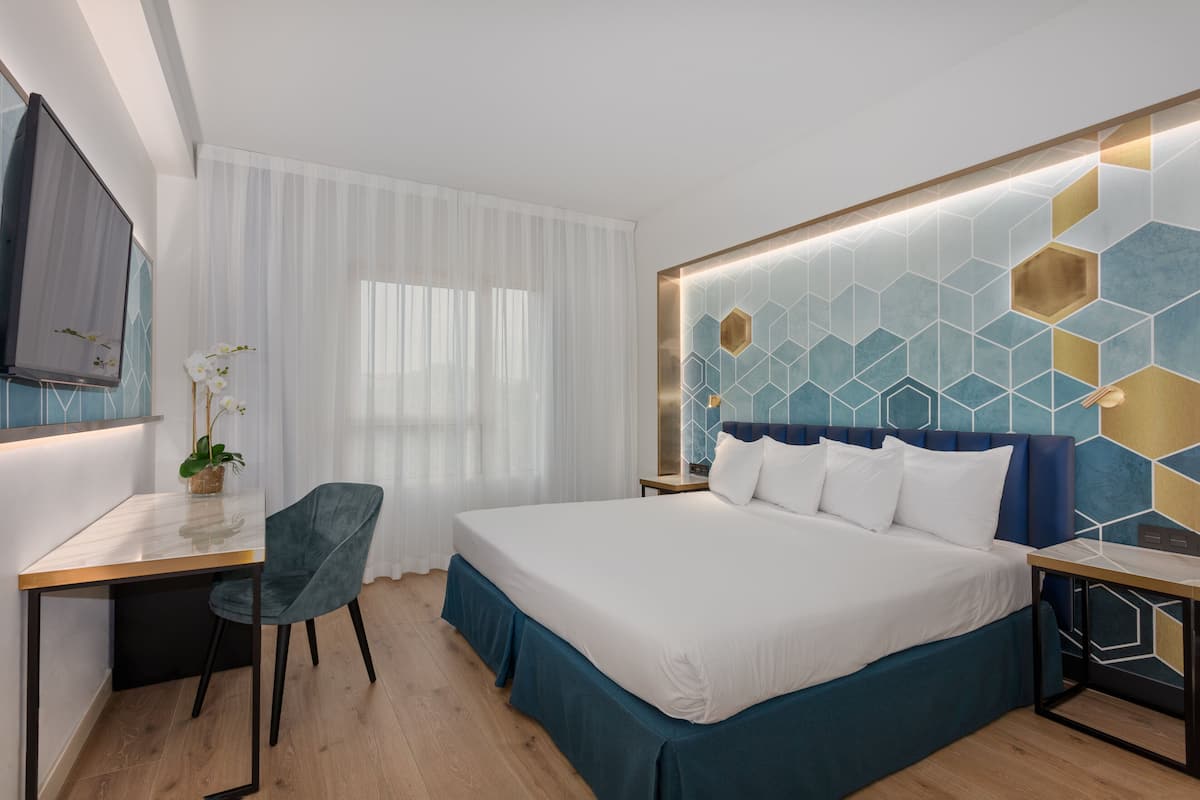 Quarto de hotel moderno com uma cama grande, uma escrivaninha com cadeira, uma televisão e um painel decorativo.