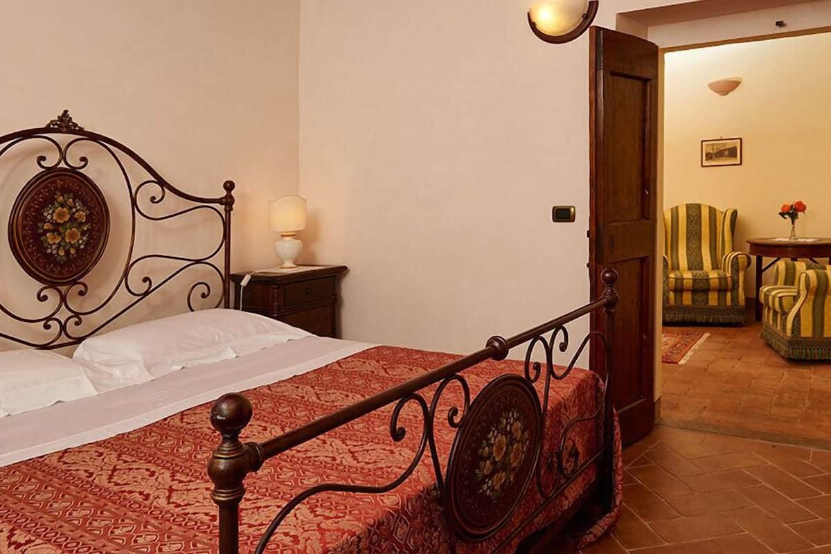 Uma cama bem arrumada com colcha estampada, uma mesa de cabeceira com abajur e uma cadeira num quarto acolhedor, com piso de azulejos e uma porta que dá para outro espaço com mais assentos.