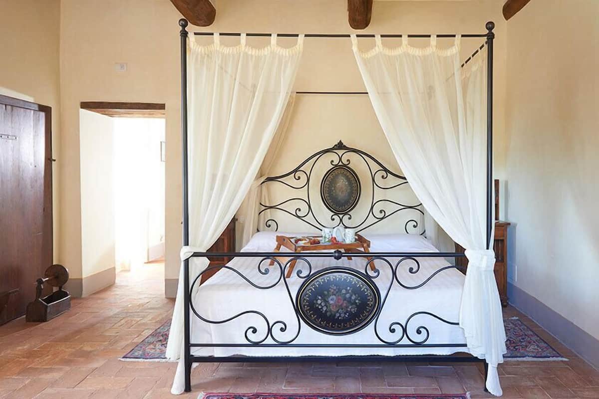 Uma cama com dossel e colunas, um pequeno criado-mudo com bandeja e um tapete estampado no chão.