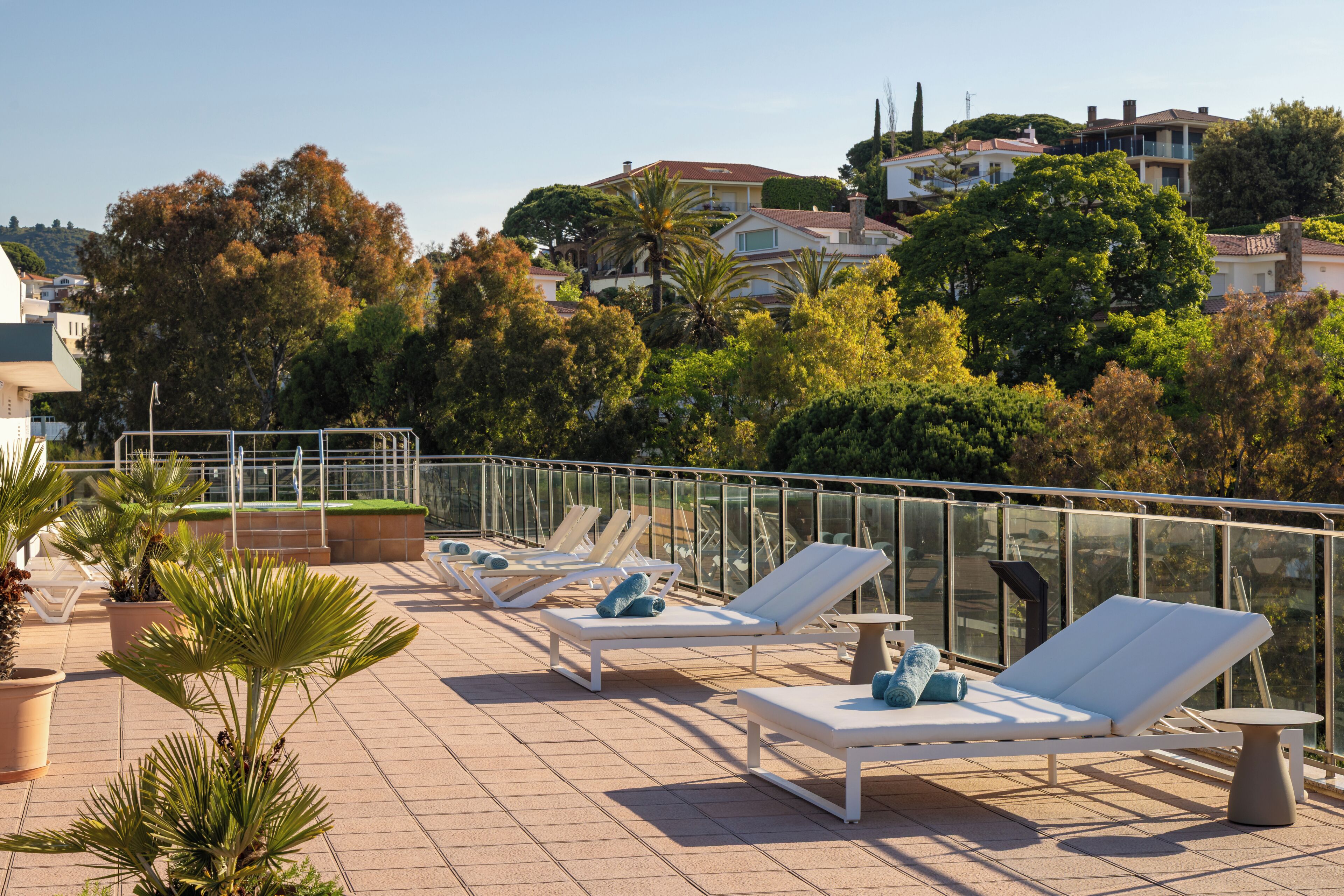 Terraço no telhado com cadeiras de lounge brancas, plantas em vasos e guarda-corpo de vidro com vista para uma área residencial.