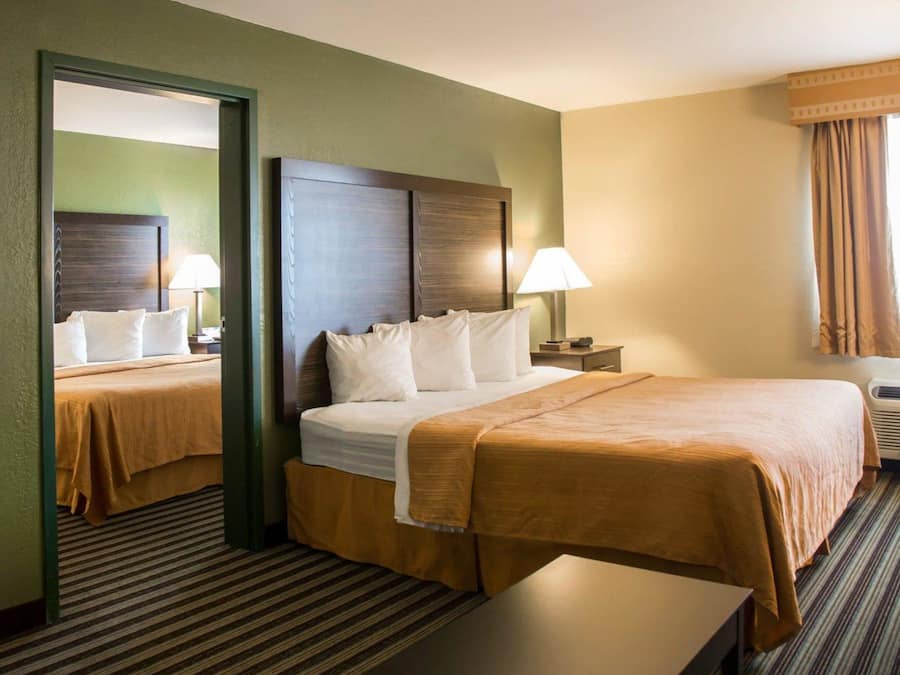 Quarto de hotel com uma cama grande, mesa de cabeceira com abajur, espelho e janela com cortinas.