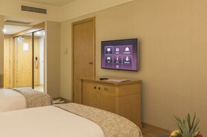 Quarto de hotel com duas camas, um armário de madeira com uma televisão mostrando opções de serviço de quarto e uma porta que dá para outro quarto.