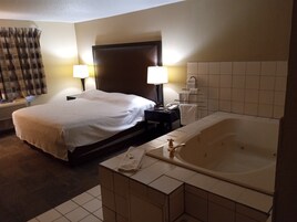 Quarto de hotel com cama, mesa de cabeceira com abajur, banheiro com banheira e janela com cortinas.