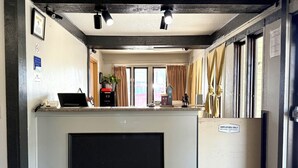 Uma mesa de recepção com um computador, um telefone e uma planta. Há placas nas paredes e uma câmera de segurança acima da mesa.