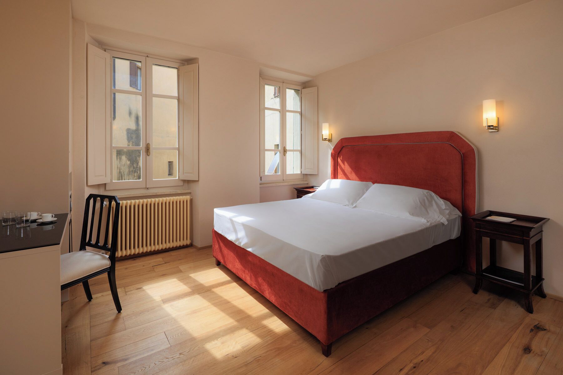 Um quarto com cama, uma cadeira, uma mesa e duas janelas.