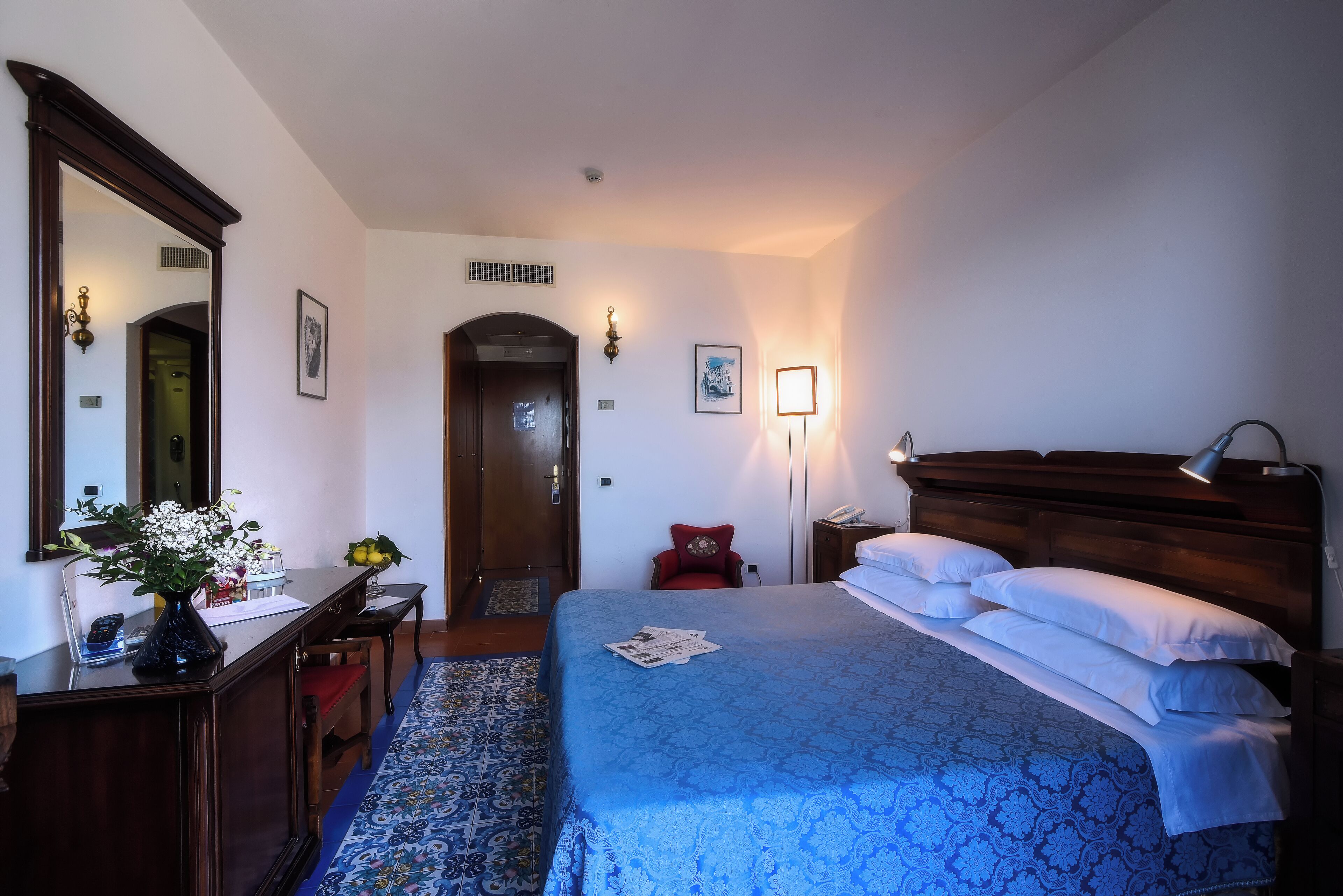 Quarto de hotel com uma cama grande, uma escrivaninha com um vaso de flores, uma cadeira, um abajur e uma porta que dá para outro cômodo.