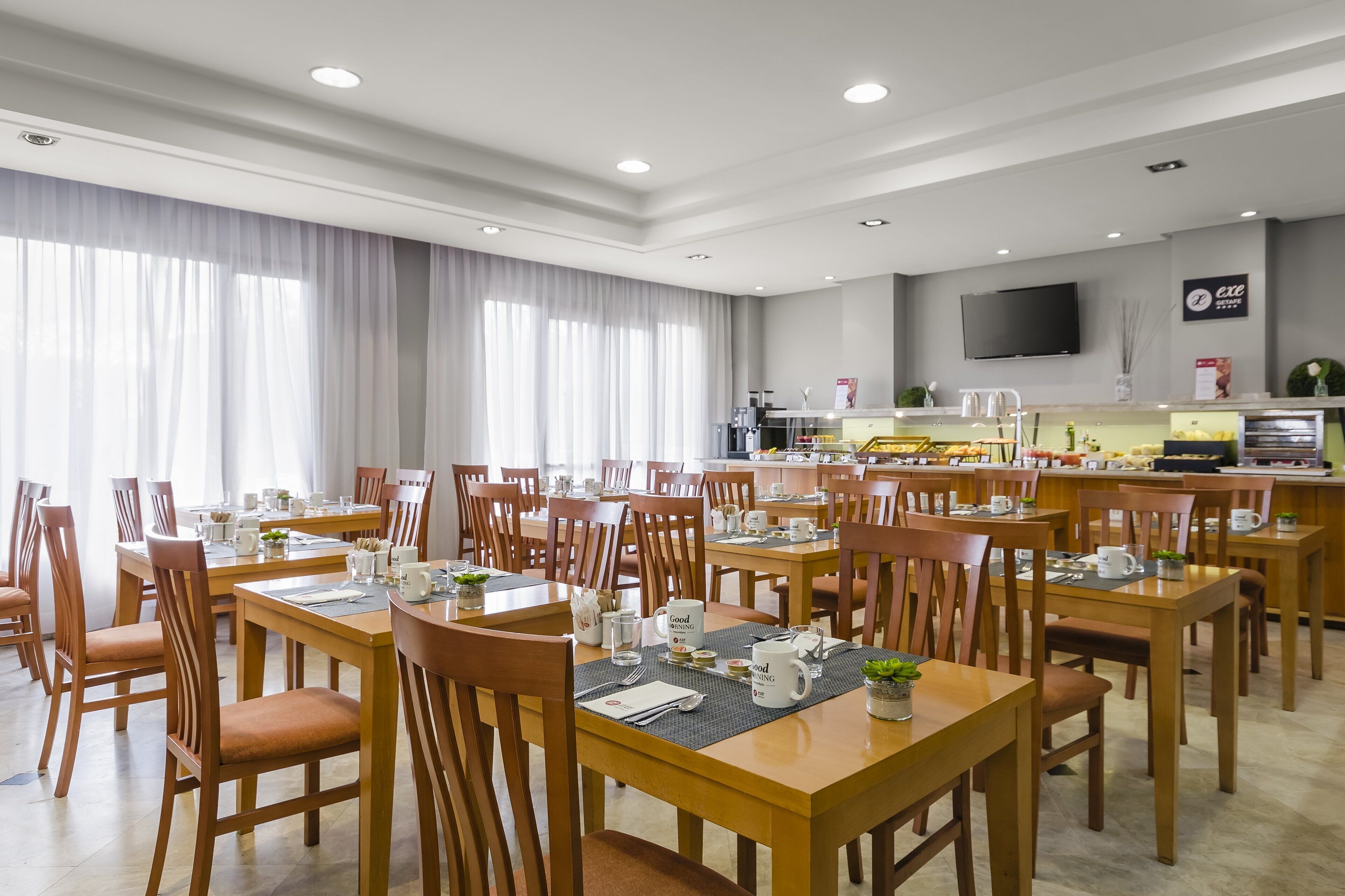 Uma sala de jantar com várias mesas e cadeiras de madeira, uma cozinha com televisão de ecrã plano e uma janela grande com cortinas.