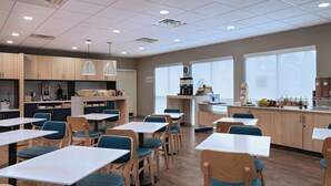 Uma sala de jantar moderna com mesas e cadeiras de madeira, uma área de cozinha com um relógio e grandes janelas que oferecem iluminação natural.